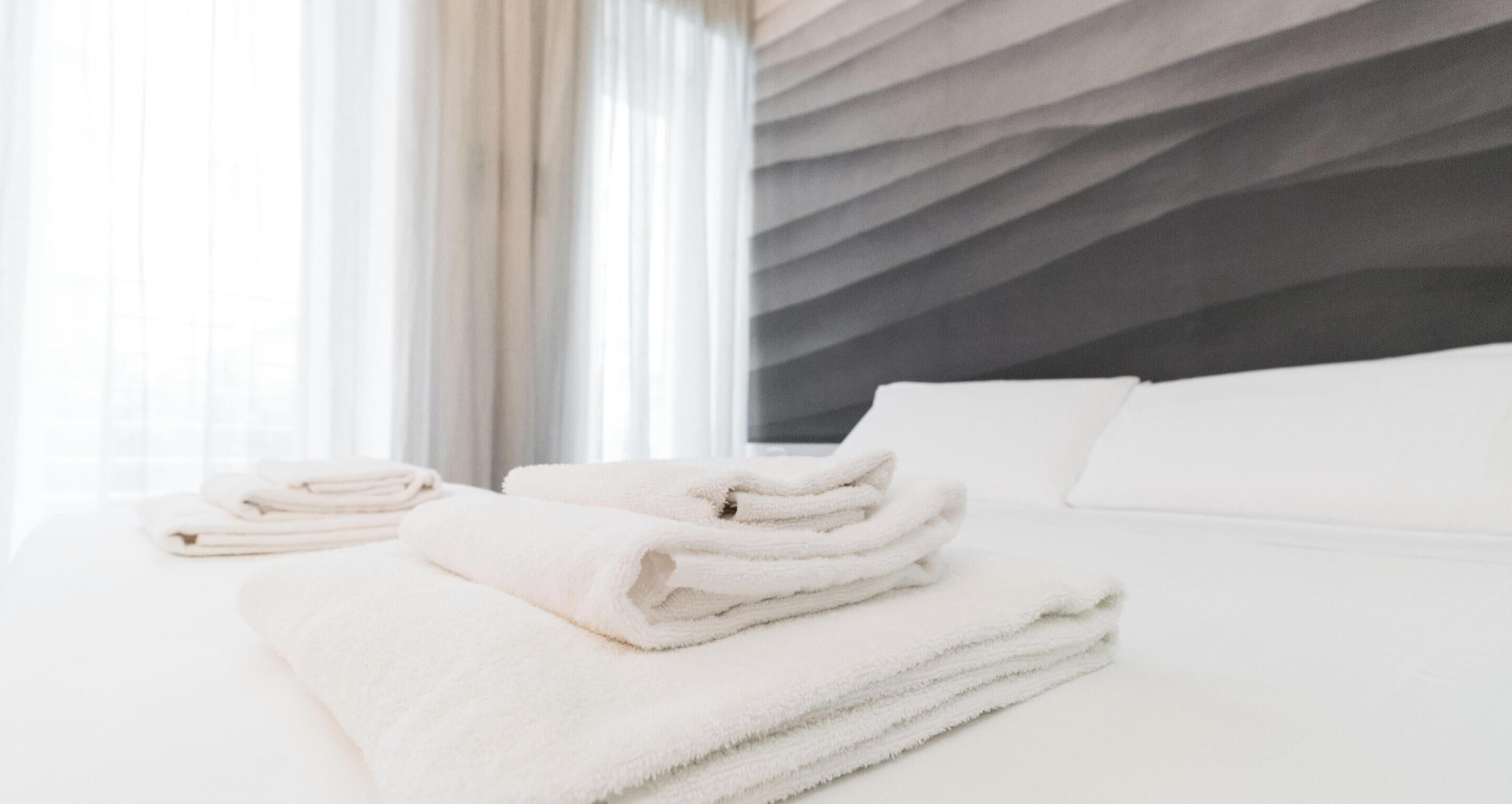 Cama bem arrumada com toalhas brancas dobradas e cabeceira moderna com textura.