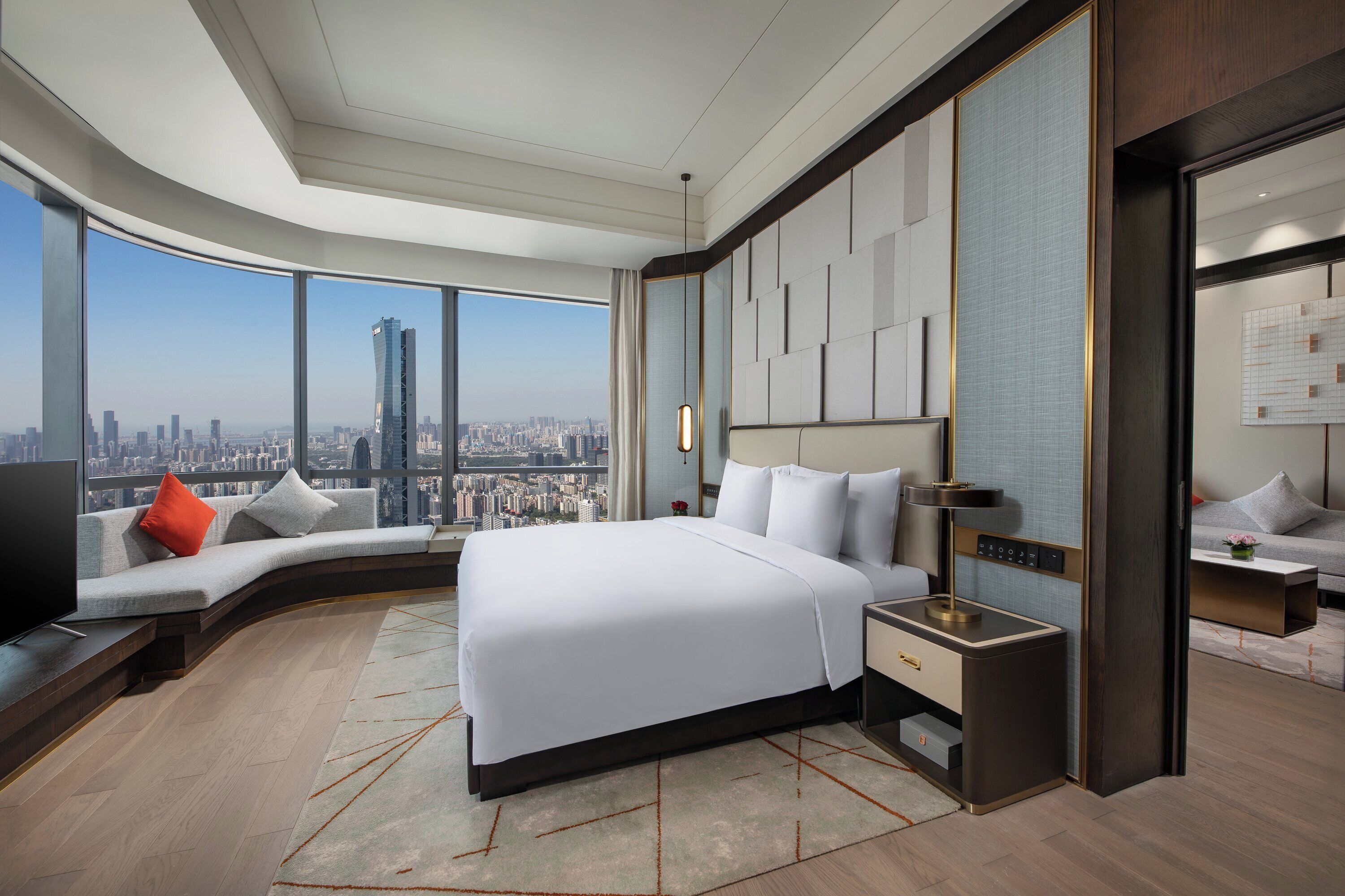 Quarto de hotel moderno com cama arredondada, uma janela ampla com vista para a cidade, um sofá com almofadas e uma mesa de cabeceira com luminária.