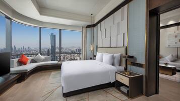 Quarto de hotel moderno com cama arredondada, uma janela ampla com vista para a cidade, um sofá com almofadas e uma mesa de cabeceira com luminária.