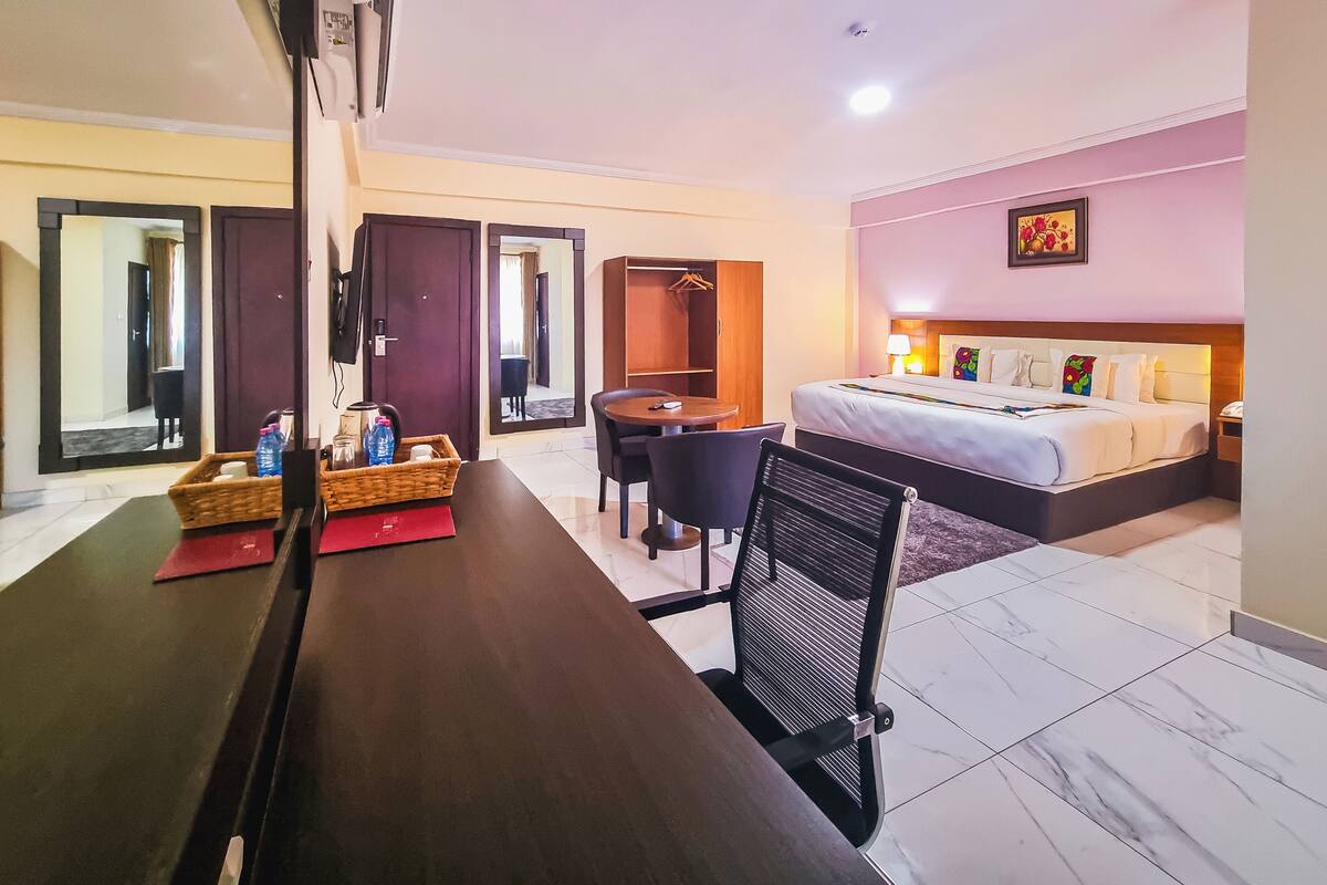 Quarto de hotel com cama, escrivaninha, cadeira, televisão, espelho e guarda-roupa.