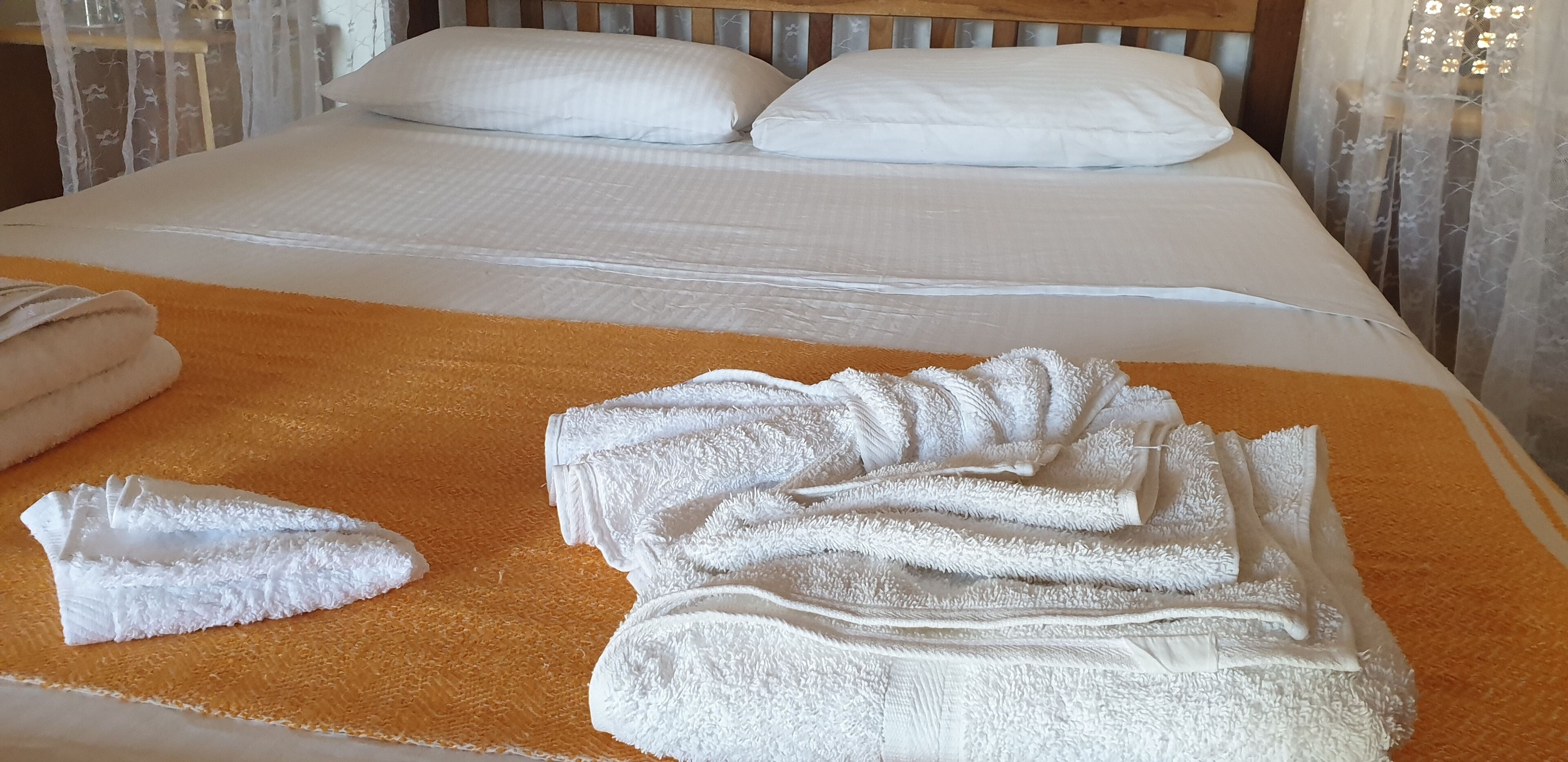Cama bem arrumada com lençóis brancos e manta laranja, acompanhada de toalhas brancas dobradas.