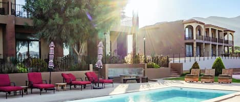 Uma área de piscina com cadeiras de sol vermelhas iluminadas e guarda-sóis, céu claro e um edifício com varandas.