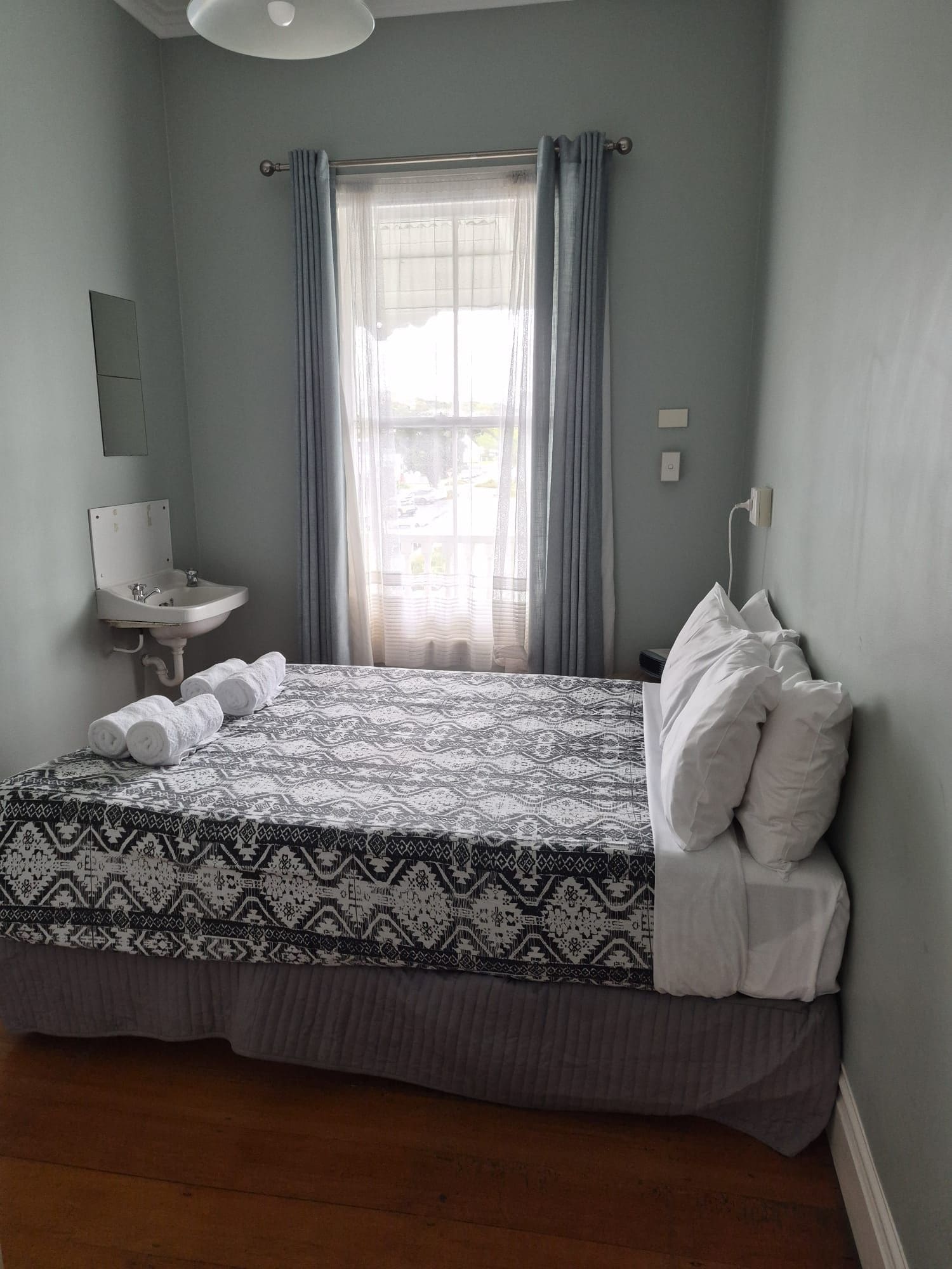 Quarto com cama, mesa de cabeceira, luminária na parede, janela com cortinas e vista para o exterior.