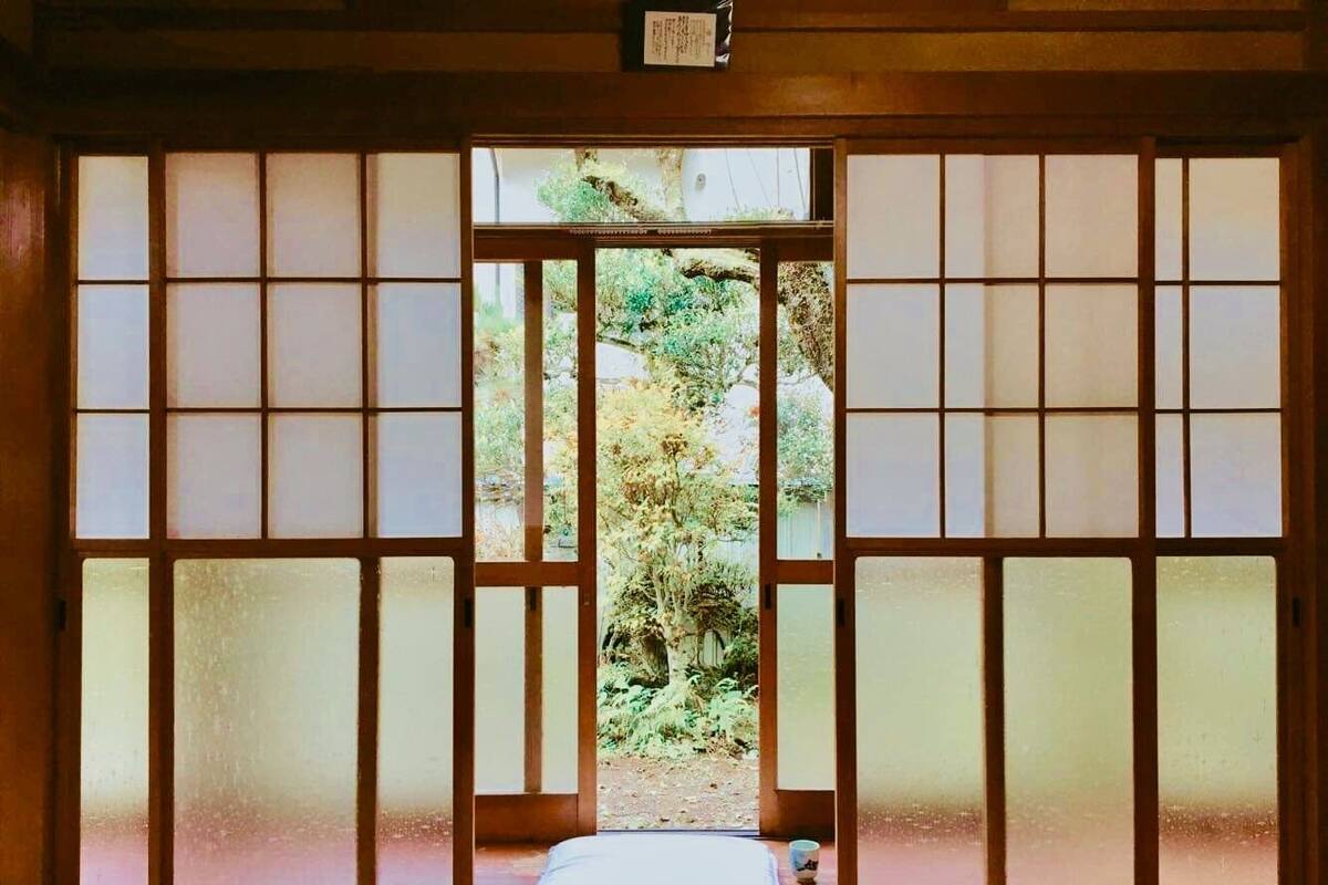 Um quarto japonês tradicional com tatames, uma cama de madeira baixa e vista para um jardim através de biombos shoji.