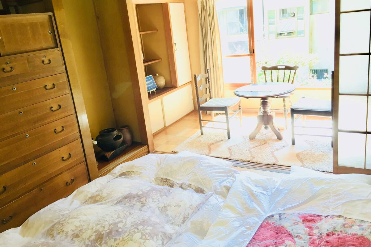 Quarto com cômoda de madeira, cama com colcha floral, mesinha com cadeira e vista para o exterior pela janela.