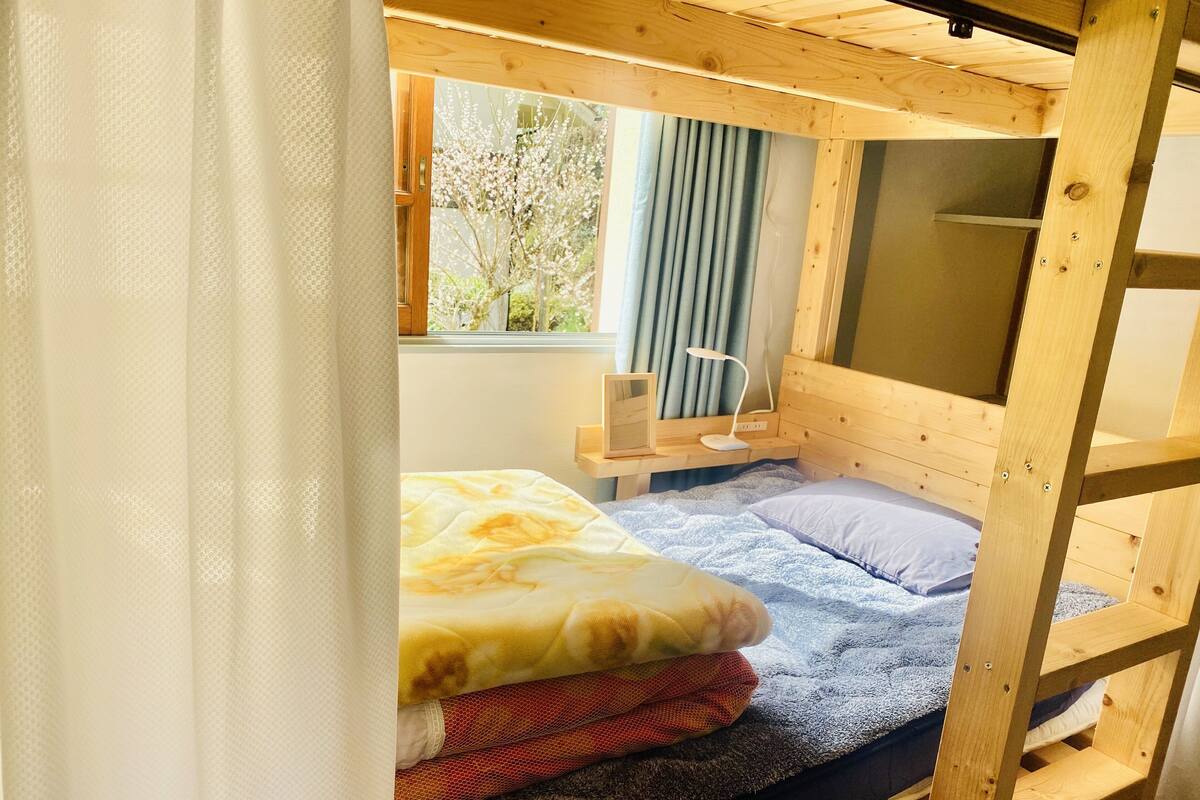 Uma cama de solteiro com estrutura de madeira, uma mesa de cabeceira com abajur e uma janela com cortinas.