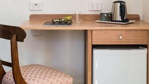 Quarto compacto com uma secretária de madeira, uma cadeira, um pequeno frigorífico, uma chaleira e uma televisão de ecrã plano na parede.