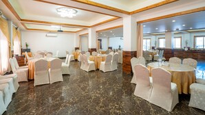 Uma ampla sala de banquetes com pé direito alto, vigas de madeira e piso de mármore polido. Mesas montadas com cadeiras brancas e cobertas por toalhas douradas.