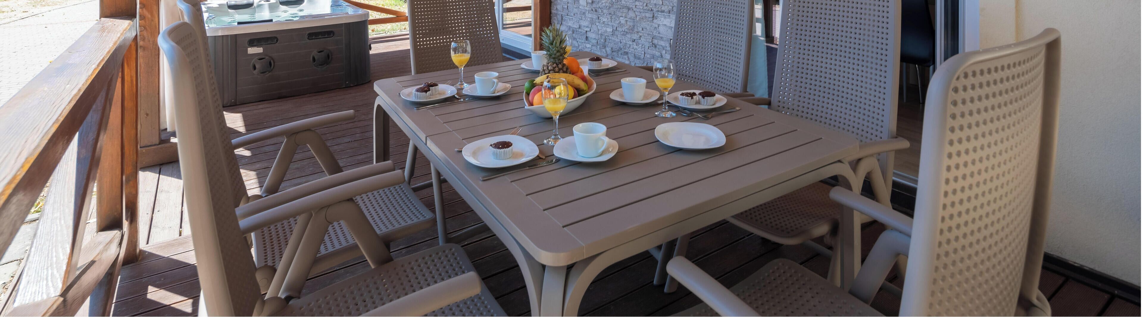 Uma área de refeições ao ar livre com uma mesa posta para dois, apresentando uma cesta de frutas, pratos, copos e uma banheira de hidromassagem ao fundo.