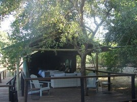 Área de estar coberta ao ar livre com sofá e cadeiras, cercada por árvores.
