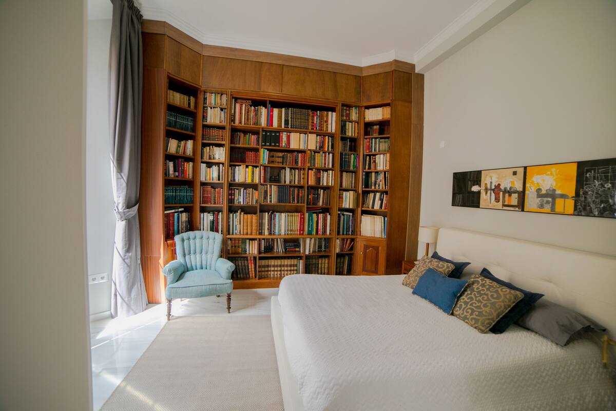 Quarto com uma grande estante de livros, uma cama com vários travesseiros e uma cadeira.