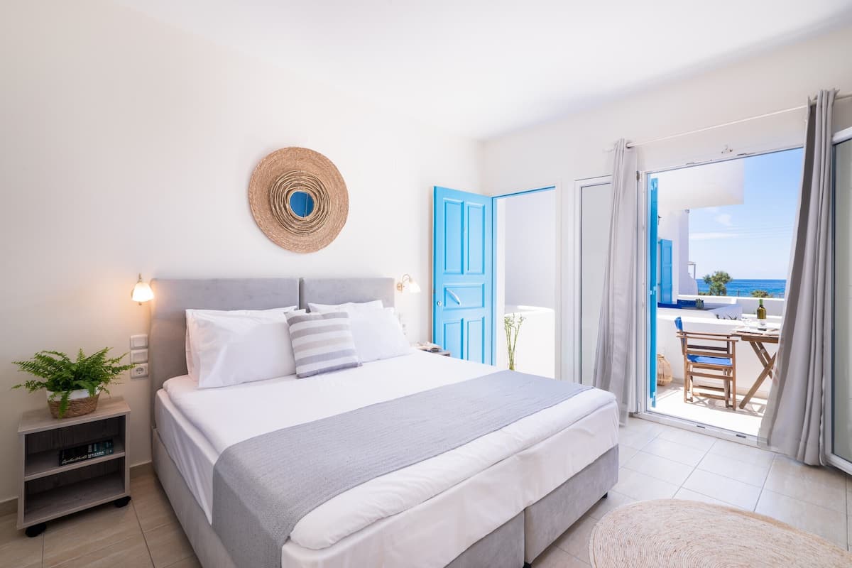 Quarto com cama, mesa de cabeceira e vista para o mar através de uma porta aberta.
