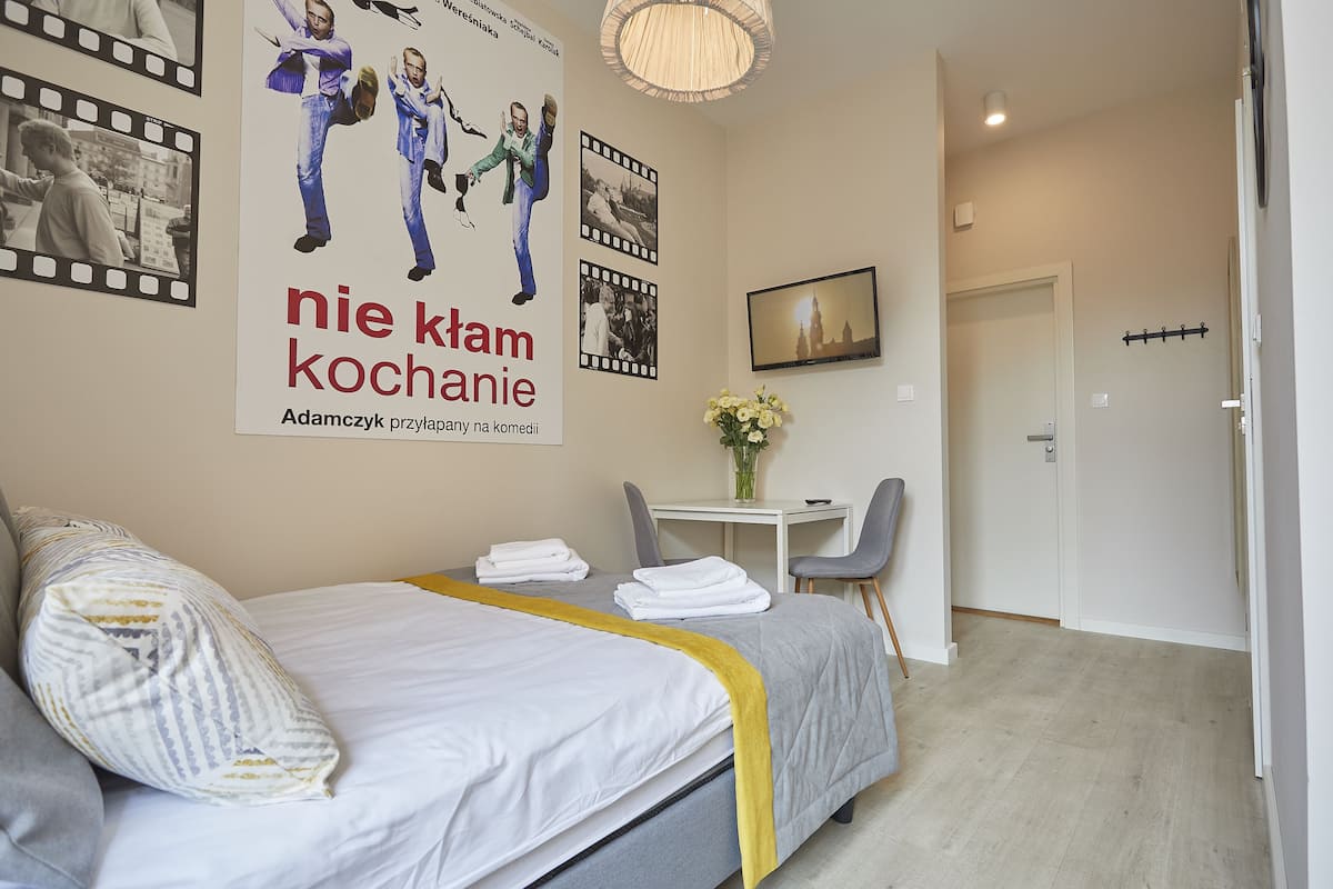 Quarto de hotel com uma cama, uma escrivaninha e uma cadeira. Há um cartaz na parede e uma porta ao fundo.
