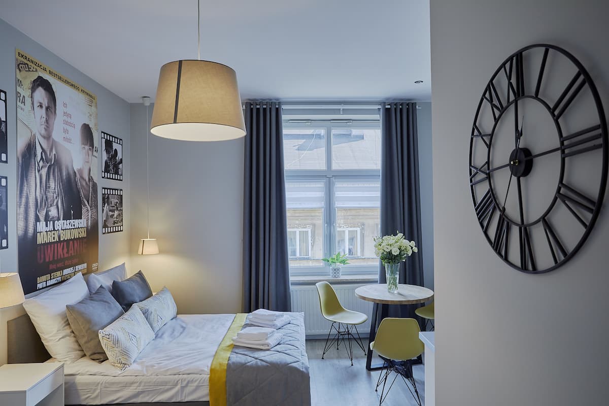 Quarto de hotel moderno com uma cama grande, um relógio redondo na parede, um espaço de refeições com uma mesa pequena e cadeiras, e uma janela com cortinas.