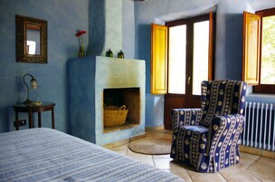 Um quarto com cama, poltrona estofada, lareira, espelho e janela com as persianas abertas.