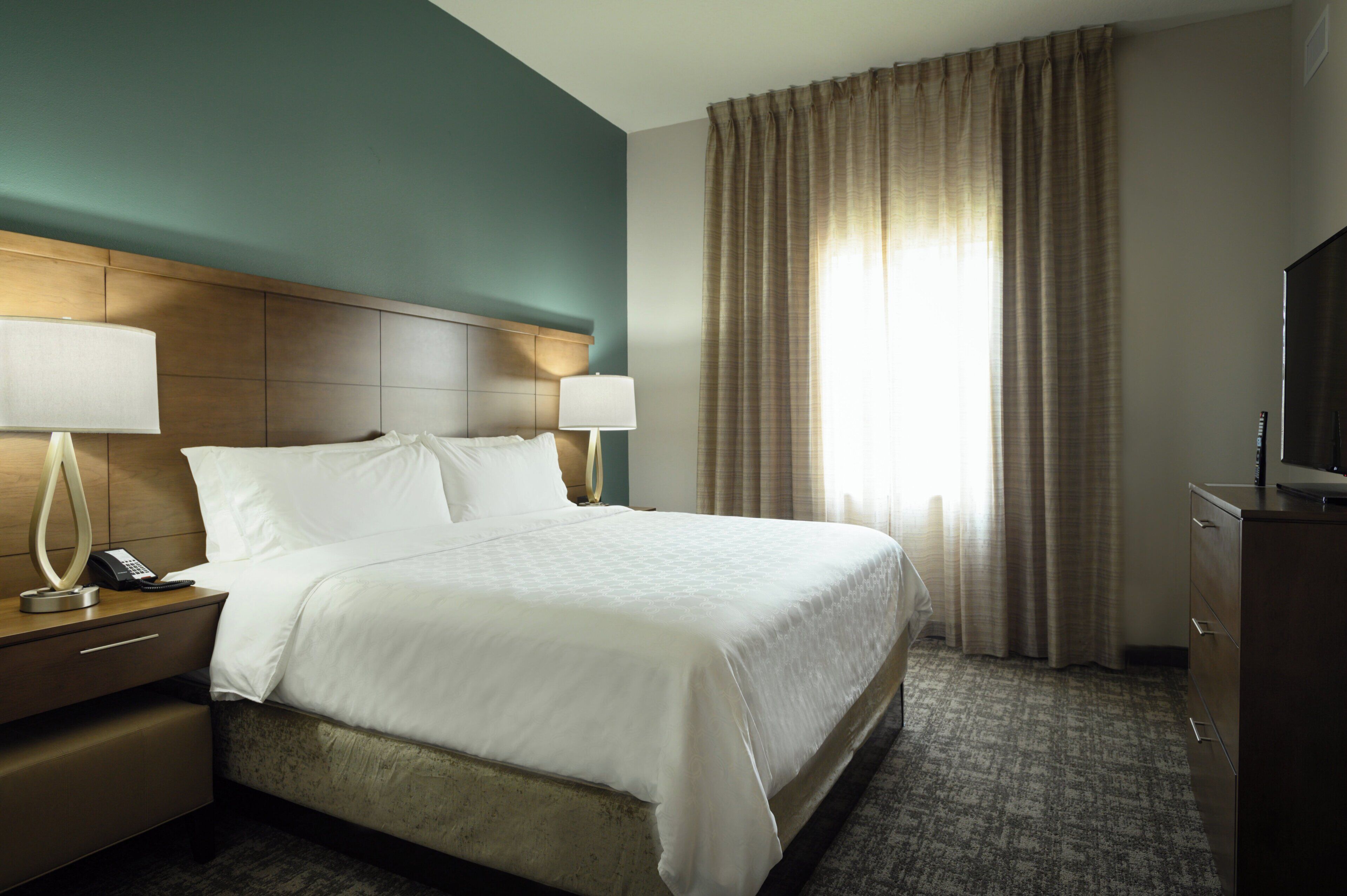 Quarto de hotel com cama, luminárias de cabeceira, telefone, escrivaninha e televisão.