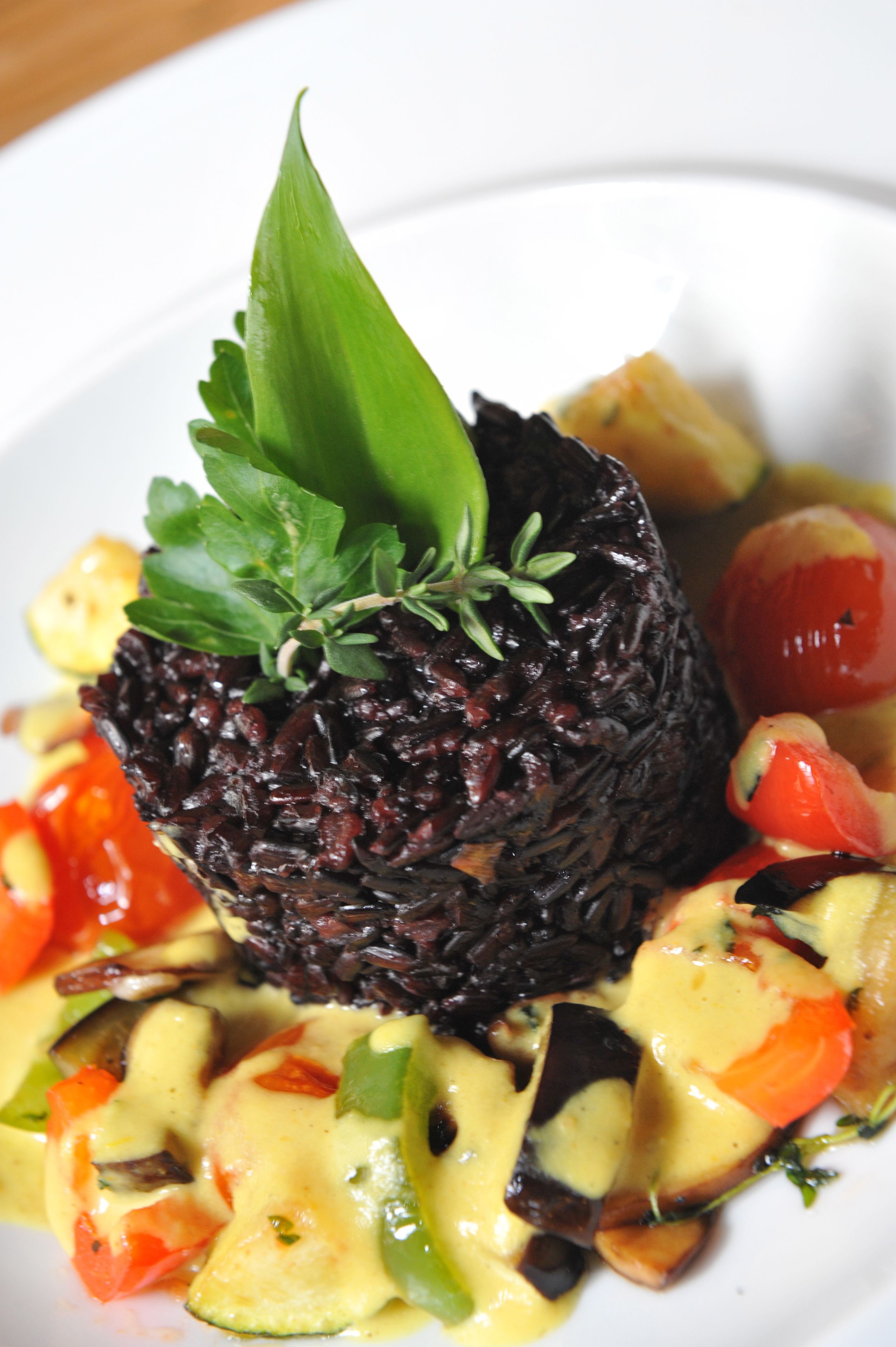 Prato com arroz preto, decorado com uma folha verde, acompanhado de legumes e molho cremoso.