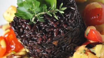 Prato com arroz preto, decorado com uma folha verde, acompanhado de legumes e molho cremoso.