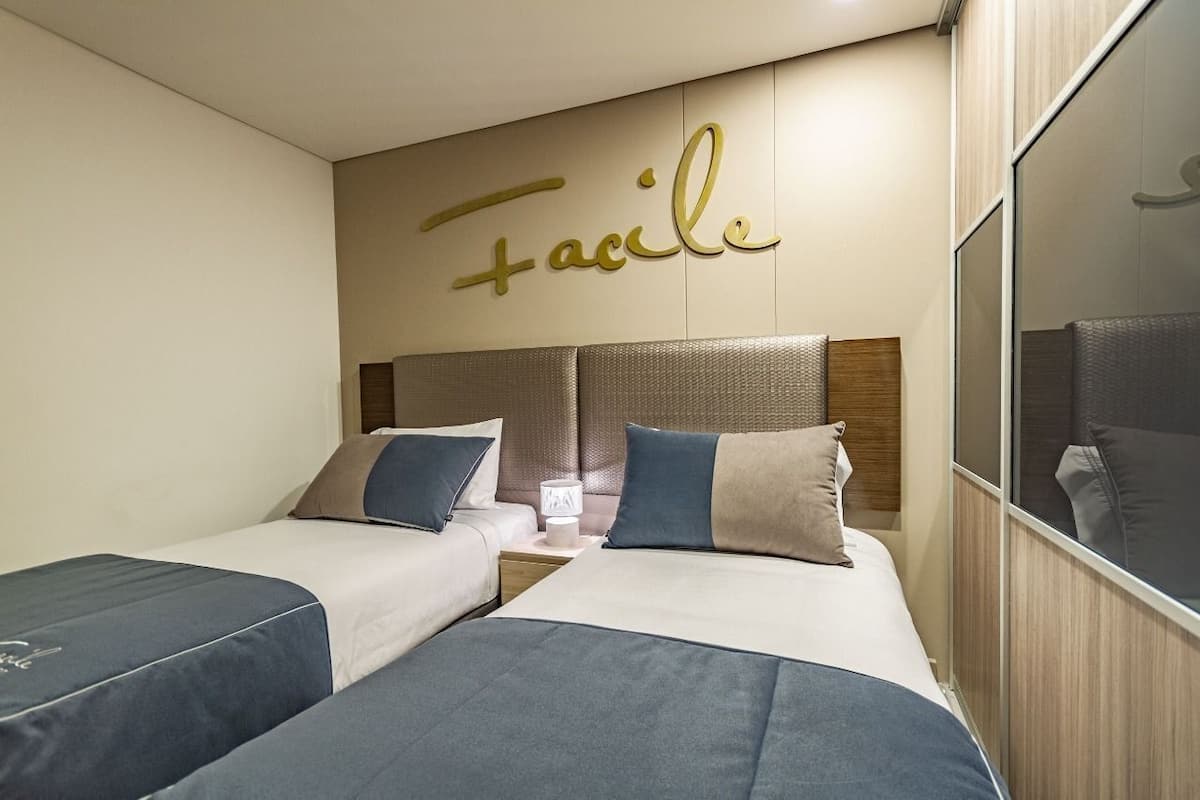Quarto de hotel com duas camas, cabeceira com a placa 'Facile', mesa de cabeceira com abajur e espelho na parede.