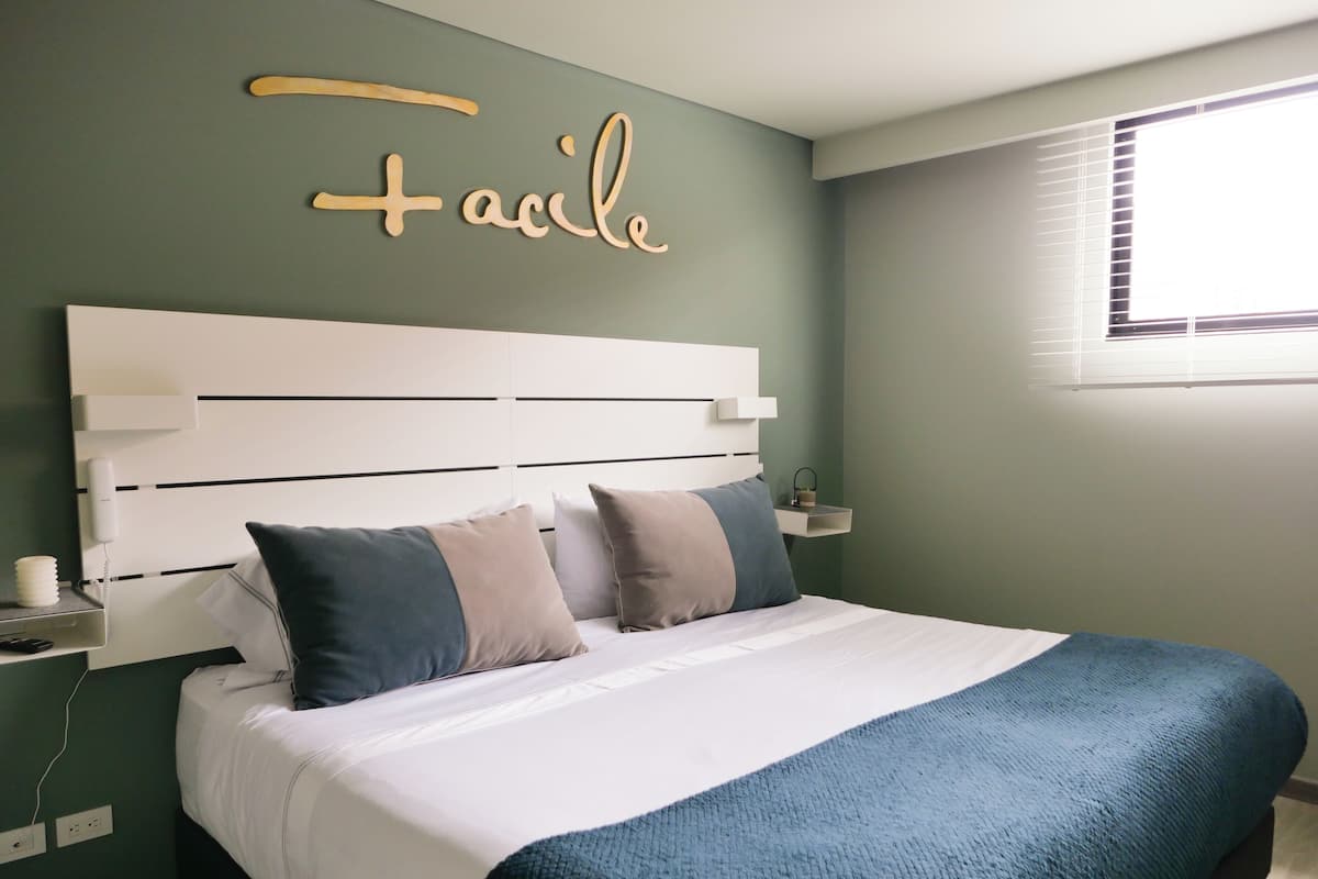 Quarto com cama, mesa de cabeceira, telefone fixado na parede e janela com persianas.