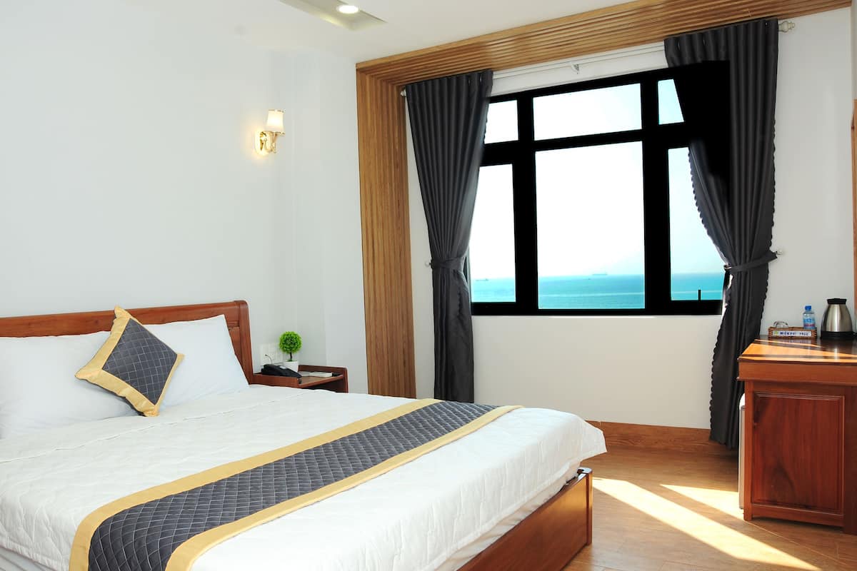 Cama bem arrumada com colcha acolchoada, mesa de cabeceira com abajur e vista para o mar através de uma janela com cortinas.