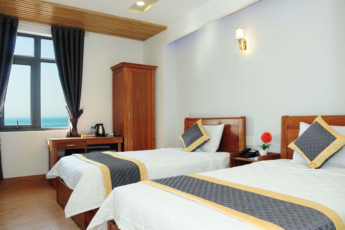 Quarto de hotel com duas camas, um guarda-roupa de madeira, uma escrivaninha, uma mesa de cabeceira e uma janela com cortinas.