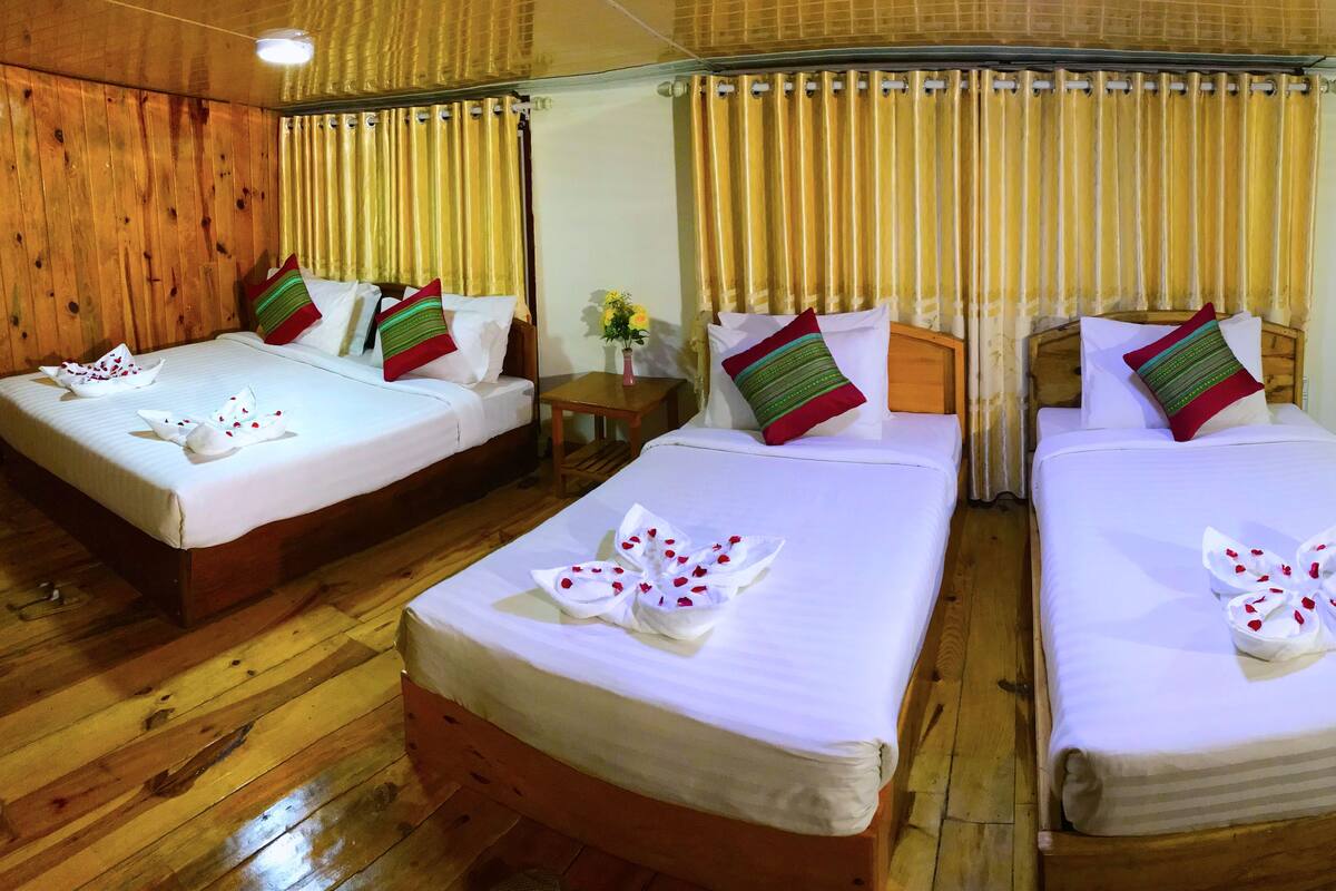 Quarto de hotel com três camas de madeira, cada uma com lençóis brancos e almofadas decorativas. Há uma pequena mesa de madeira com um vaso de flores entre as camas.