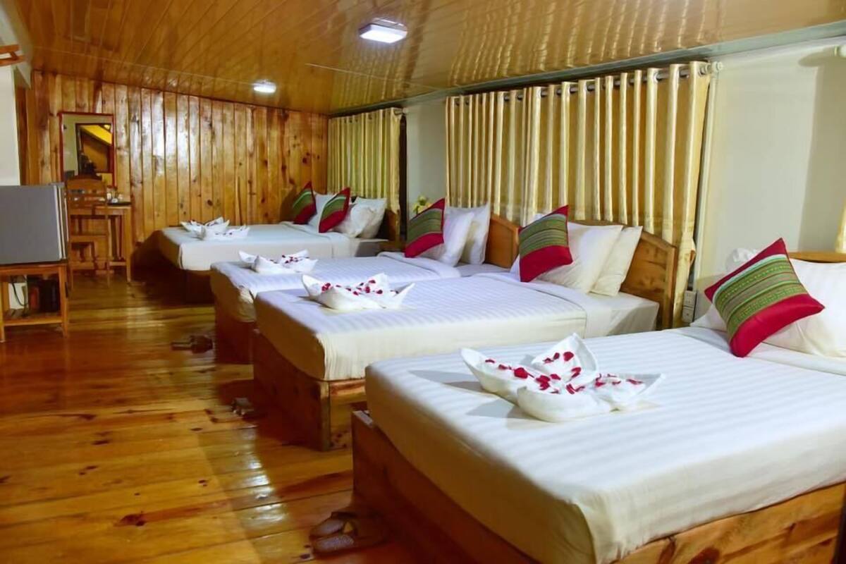 Quarto de hotel com piso de madeira, várias camas com roupa de cama branca e almofadas coloridas, cabeceira de madeira e uma pequena escrivaninha com cadeira.
