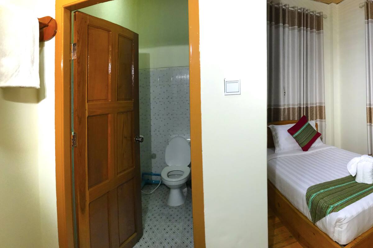 Um quarto com porta de madeira, um vaso sanitário, uma cama com manta verde, uma mesa com uma chávena e uma garrafa, e uma cadeira.
