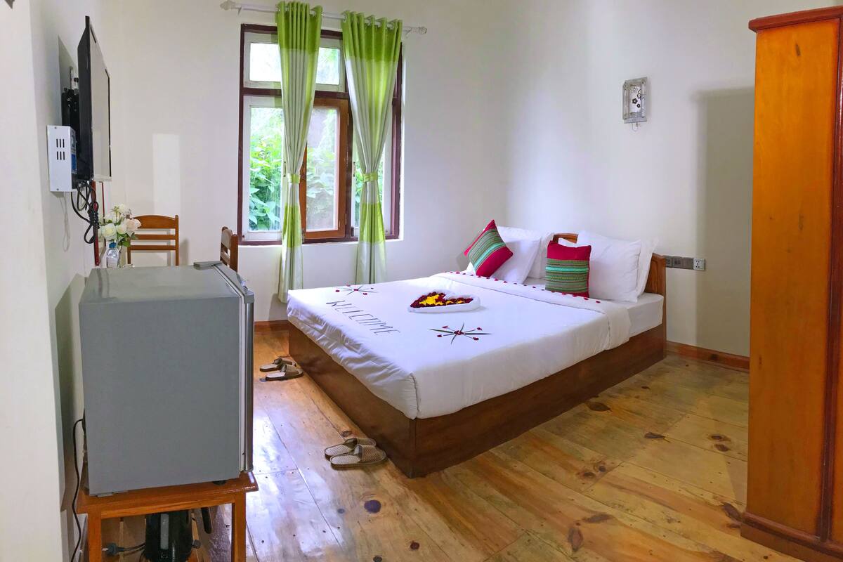 Quarto com cama, televisão sobre suporte, janela com cortinas e piso de madeira.