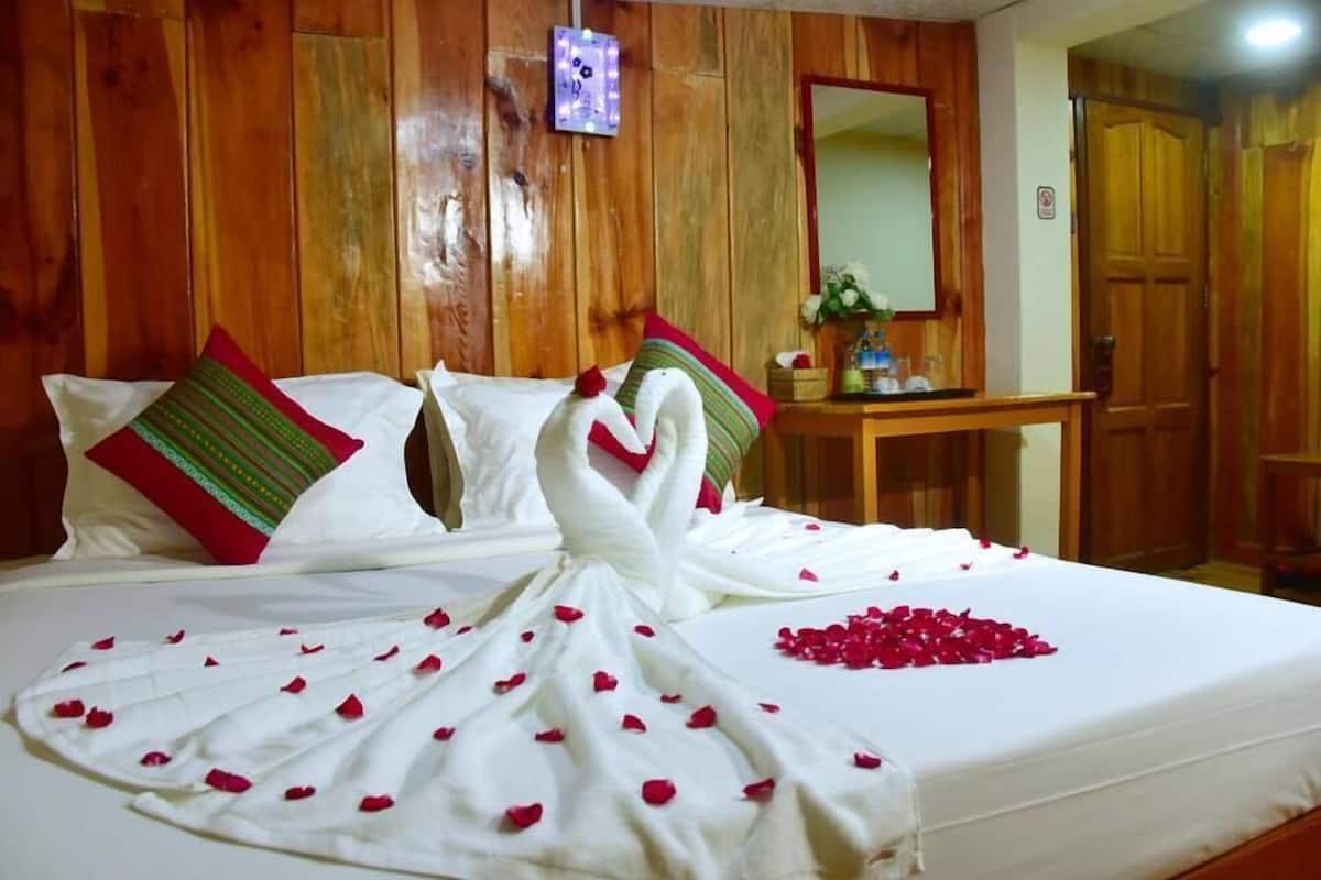 Uma cama bem arrumada com toalha em formato de cisne e pétalas de rosa, painéis de madeira nas paredes, um espelho e uma porta de madeira.