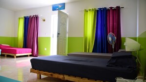 Quarto com duas camas, uma com estrutura de madeira e outra de metal. Há um ventilador na parede e uma porta que dá para outro quarto.