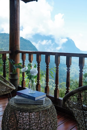 Uma cadeira de vime, uma mesinha redonda com um vaso e livros, e um guarda-corpo de madeira com vista para uma paisagem montanhosa.