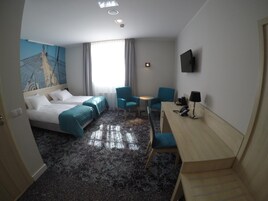 Quarto de hotel com duas camas, uma escrivaninha, uma cadeira, uma mesinha, uma televisão e um quadro na parede.
