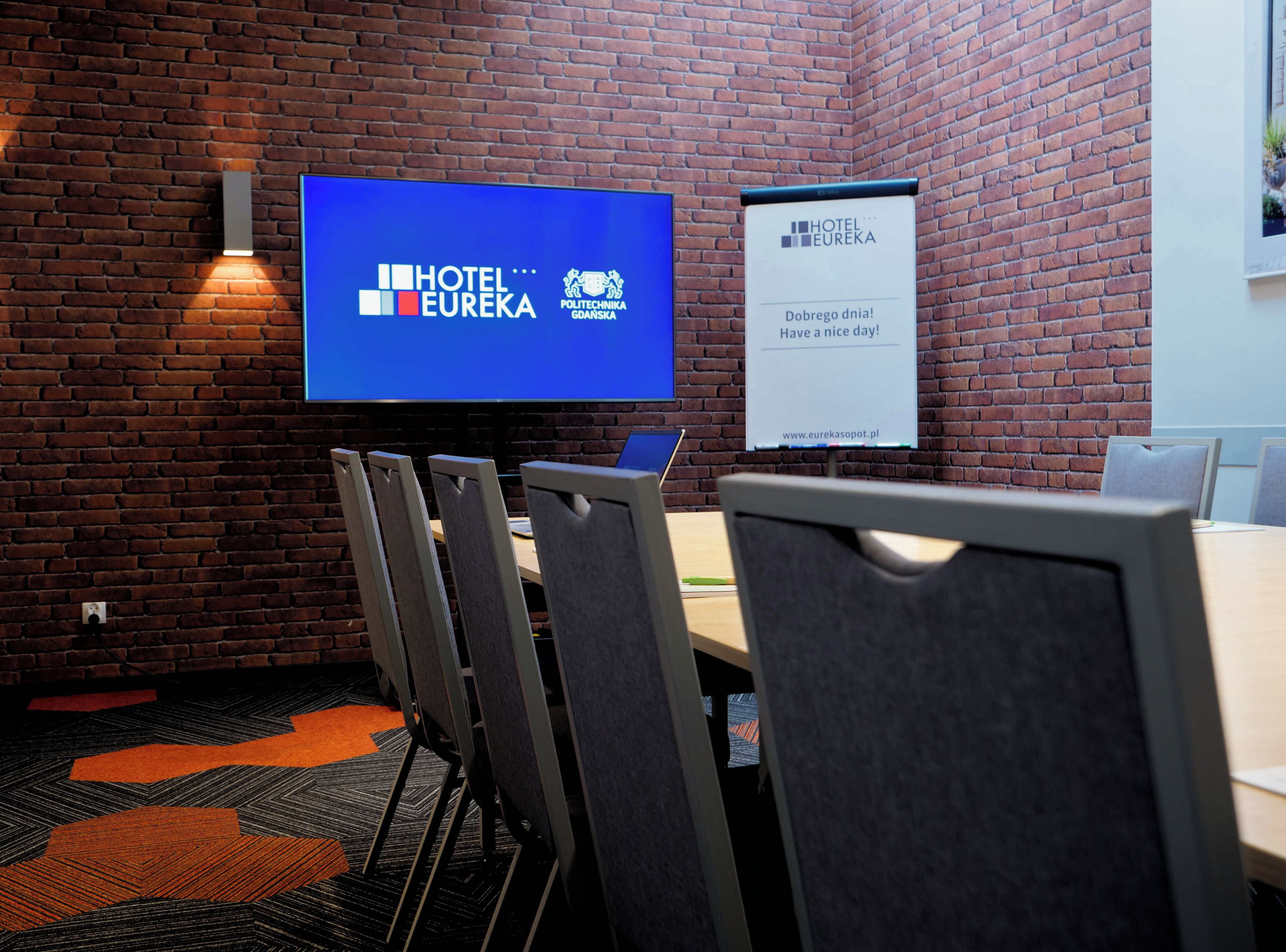 Uma sala de conferência com parede de tijolos, uma tela grande exibindo 'HOTEL EUREKA' e um letreiro com a mensagem 'HOTEL EUREKA Tenha um bom dia!'.
