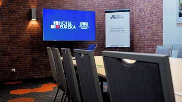Uma sala de conferência com parede de tijolos, uma tela grande exibindo 'HOTEL EUREKA' e um letreiro com a mensagem 'HOTEL EUREKA Tenha um bom dia!'.