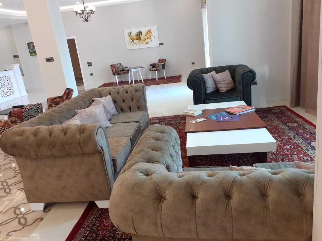 Uma sala de estar ampla com um sofá bege grande, uma poltrona menor e uma mesa de centro. Há um tapete vermelho estampado no chão e uma pintura na parede.