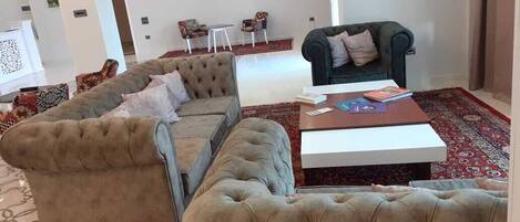 Uma sala de estar ampla com um sofá bege grande, uma poltrona menor e uma mesa de centro. Há um tapete vermelho estampado no chão e uma pintura na parede.
