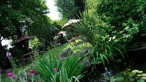 Um jardim com lago, rodeado por vegetação exuberante e um deck de madeira com guarda-sóis.
