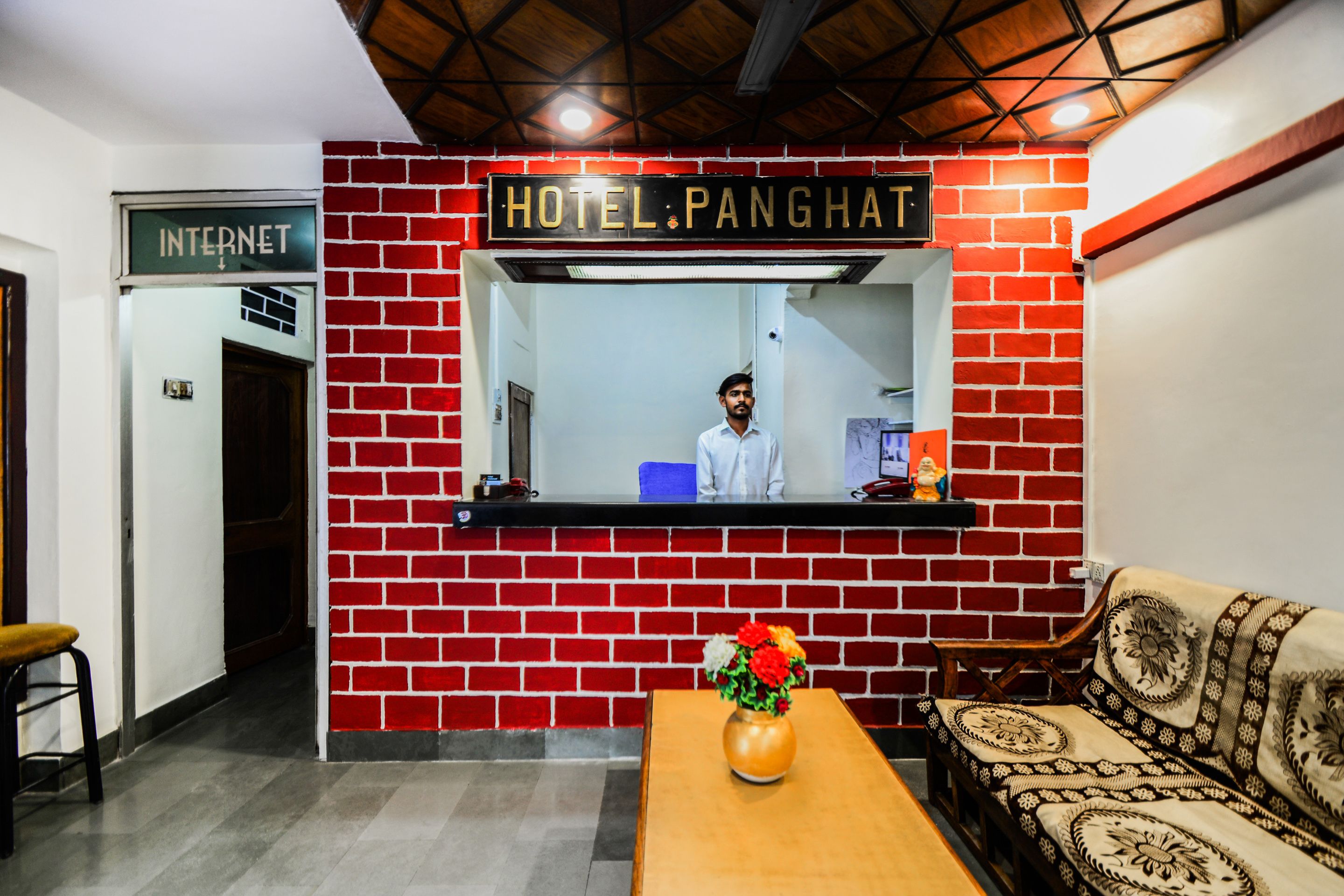 Uma área de recepção com parede de tijolos vermelhos, uma placa com a inscrição "HOTEL PANGHAT", um balcão de recepção, um sofá e um vaso com flores.