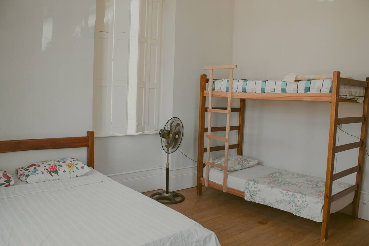 Quarto com cama de solteiro, beliche e ventilador.
