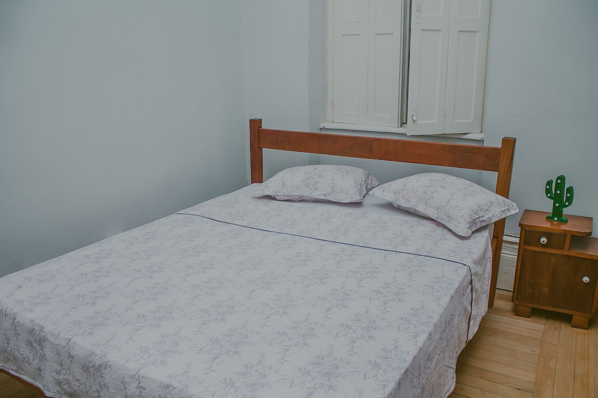 Uma cama de solteiro com cabeceira de madeira, uma mesa de cabeceira com um cacto e uma janela com persianas brancas.