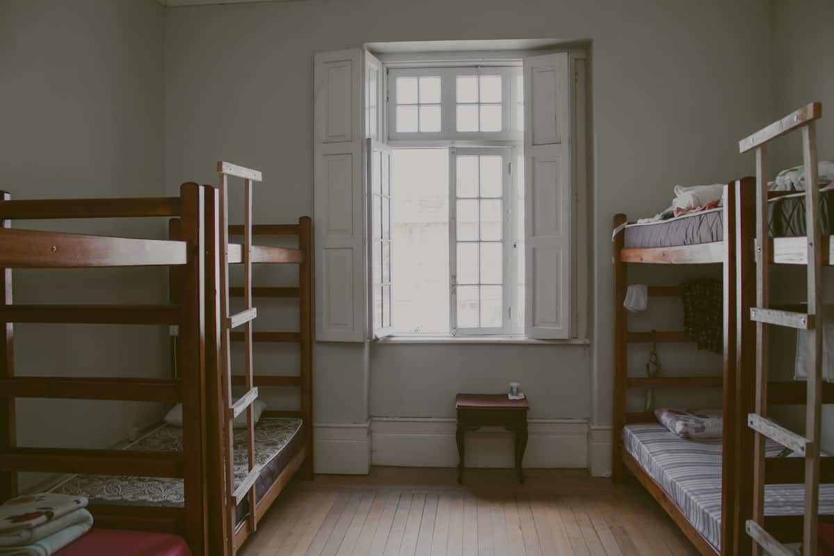 Quarto de estudante com duas beliches, uma mesa pequena e uma janela com persianas.