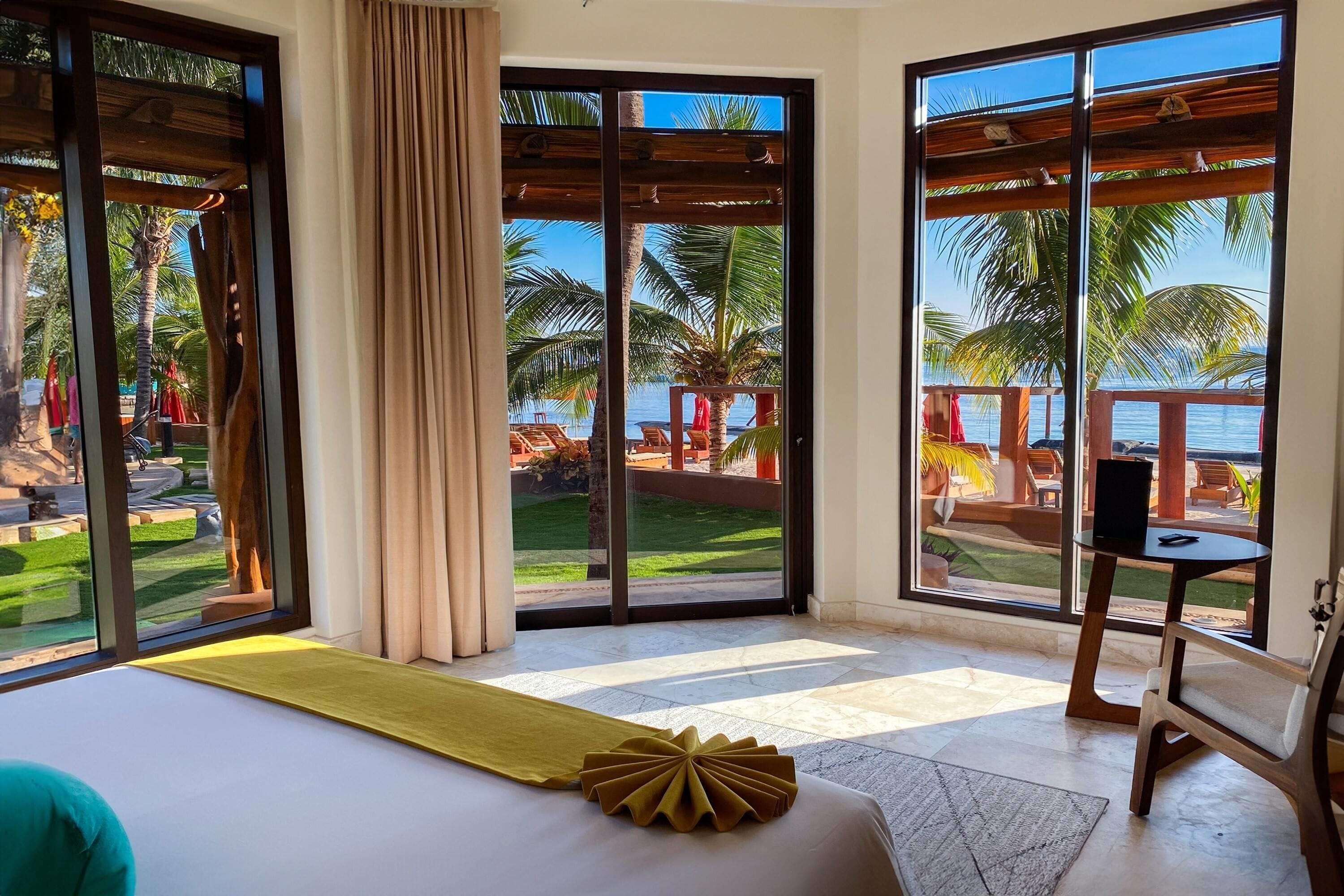 Quarto de hotel com uma grande porta de vidro deslizante para uma área da piscina, uma cama com travesseiro decorativo e uma pequena mesa com uma cadeira.