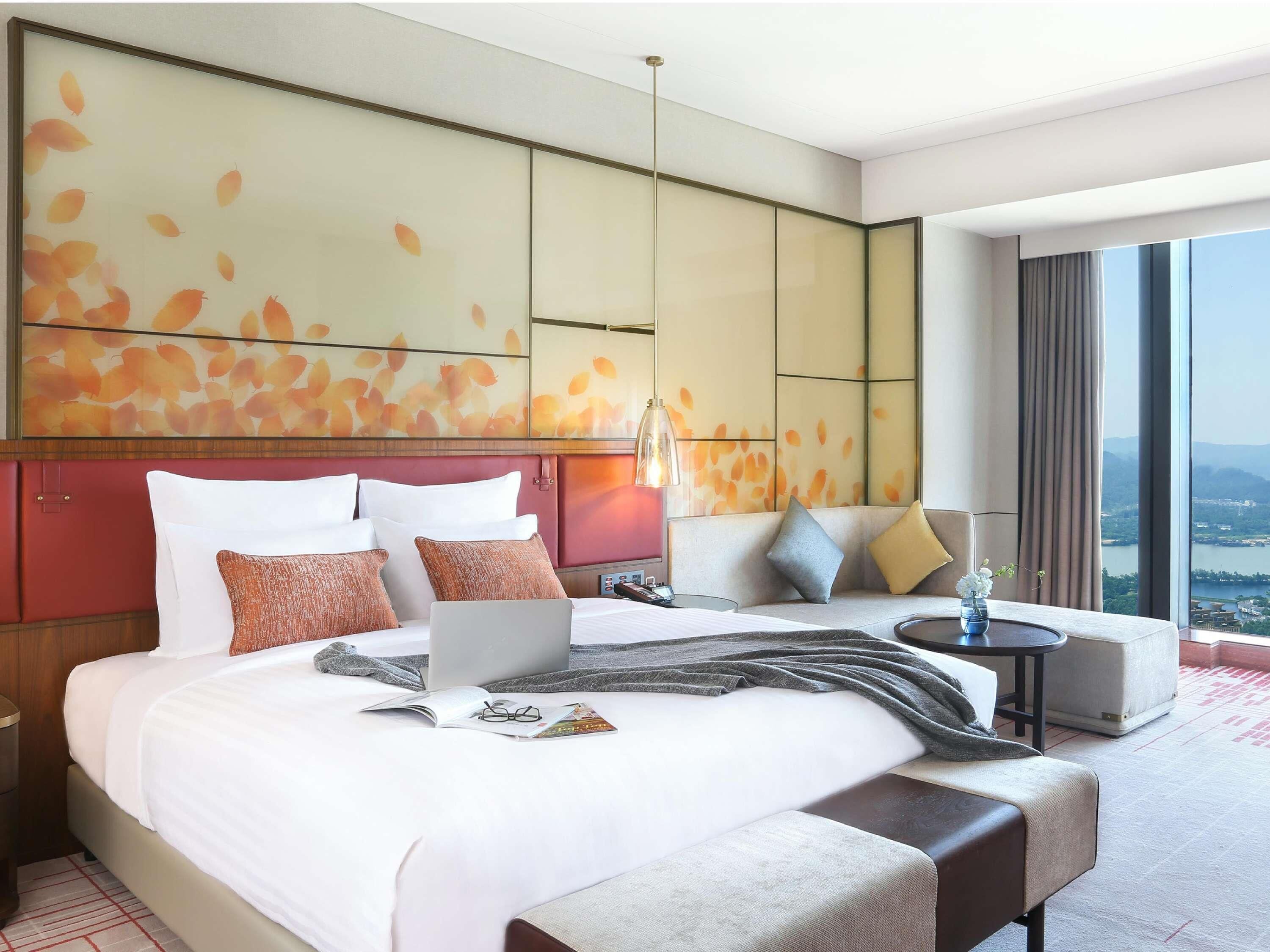 Quarto de hotel moderno com uma cama grande, área de estar e vista para um lago.