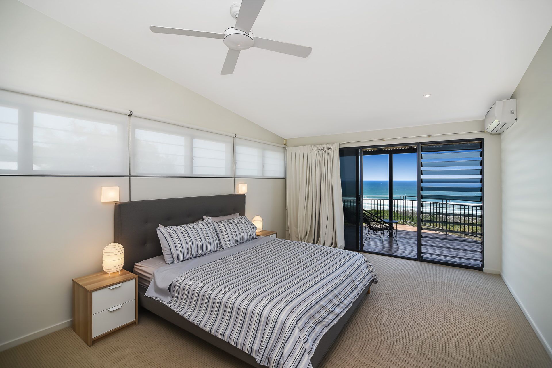 Quarto com cama, mesas de cabeceira, ventilador de teto, janela grande com cortinas e vista para o mar.