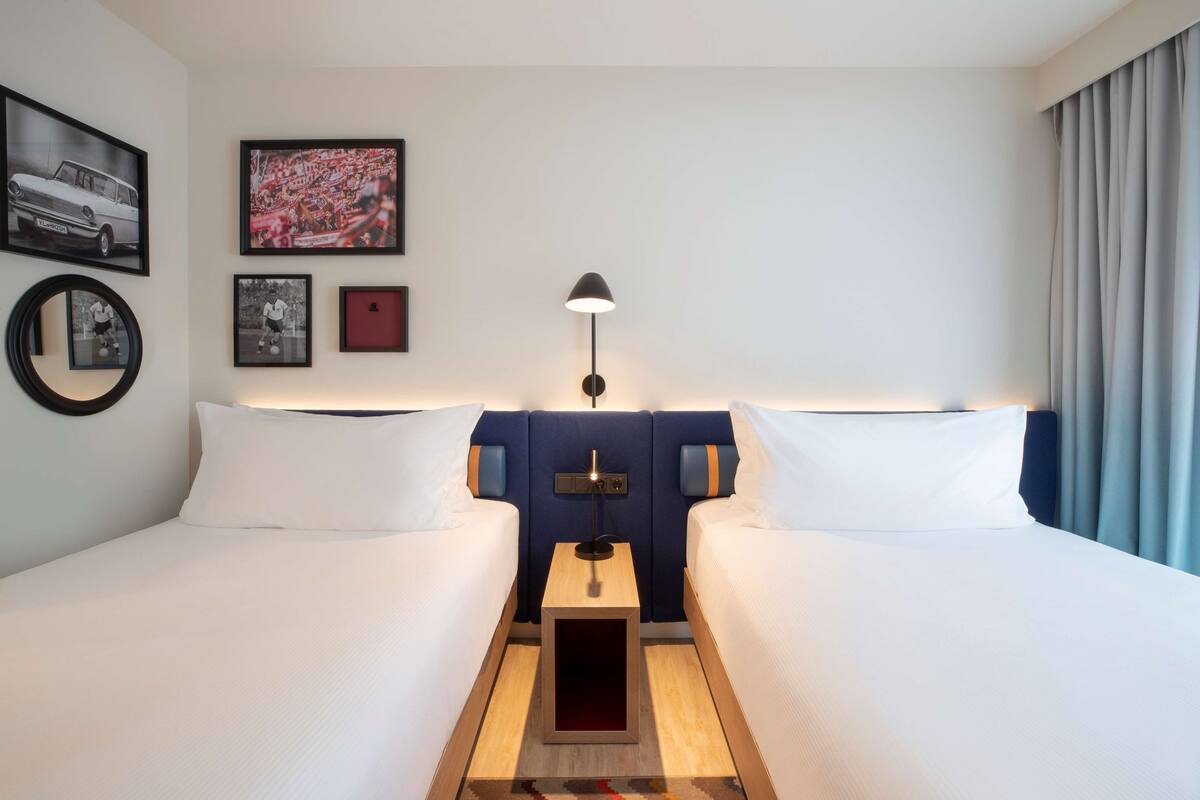 Quarto de hotel com duas camas, uma mesa de cabeceira com abajur e quadros nas paredes.