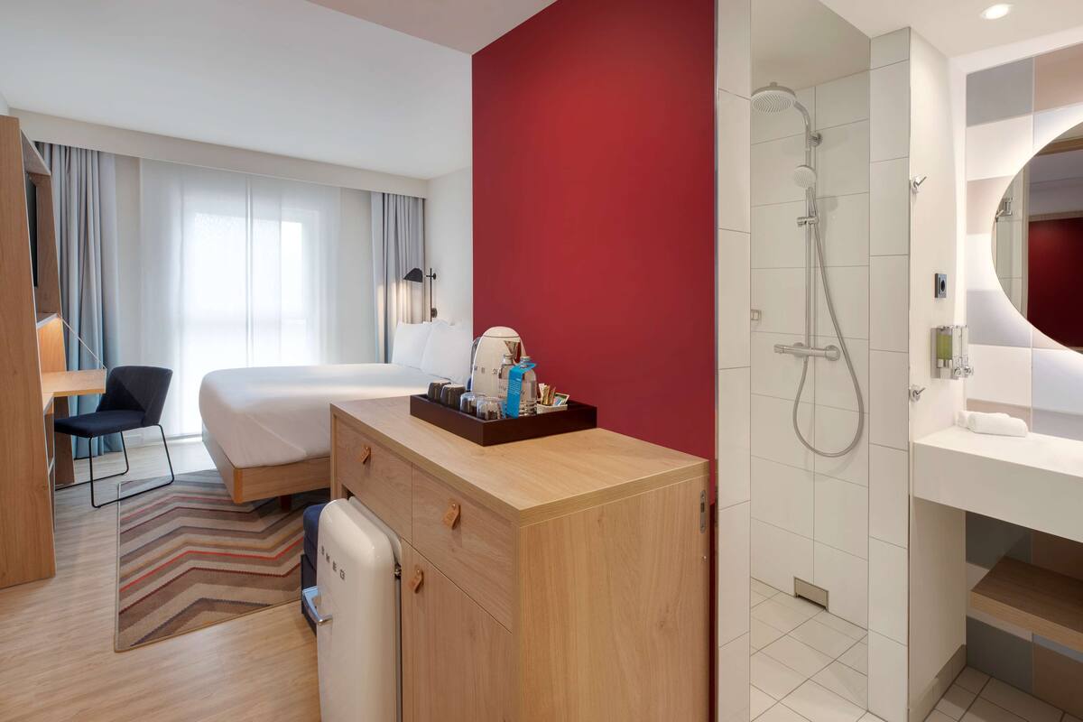 Quarto de hotel com cama, cómoda de madeira, secretária com cadeira, zona de duche e um espelho grande.
