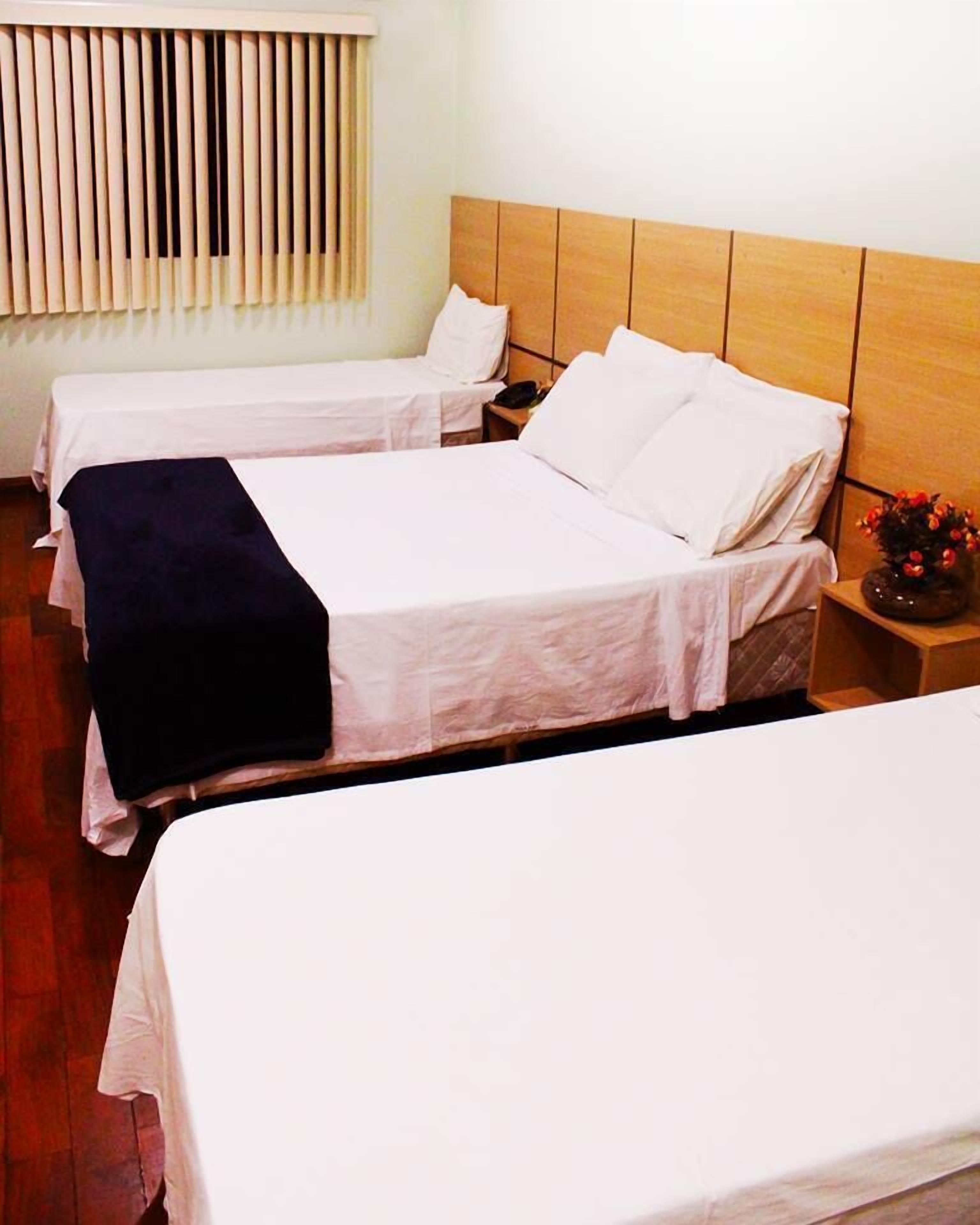 Quarto de hotel com duas camas de solteiro, cabeceira de madeira, mesa de cabeceira com um vaso de flores e janela com persianas.