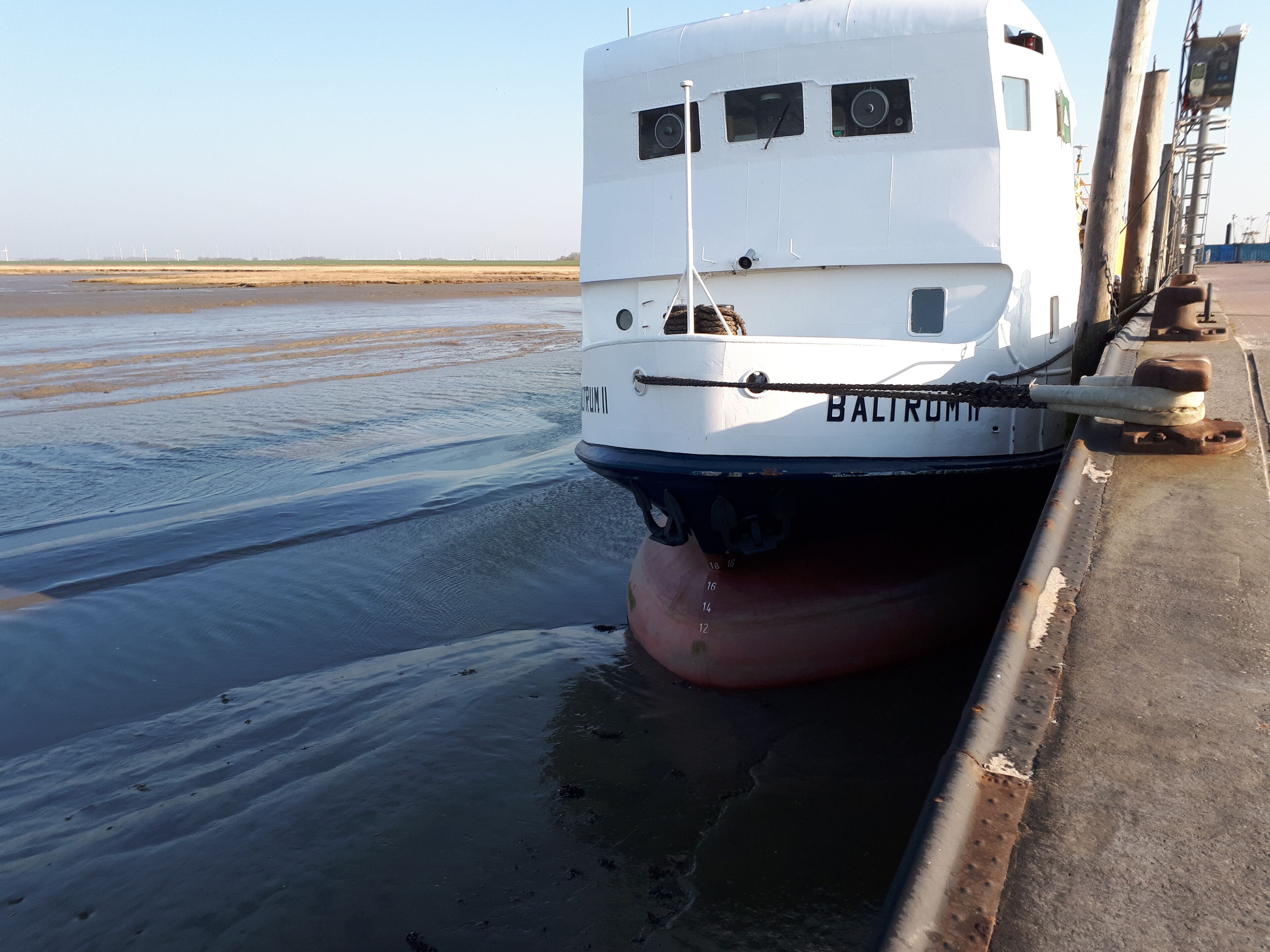Um barco chamado 'BALIRUM II' atracado em um porto com uma praia arenosa e turbinas eólicas ao fundo.